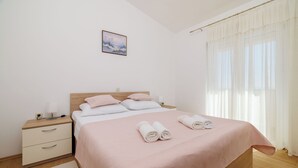 Uma cama bem arrumada com roupa de cama rosa e toalhas enroladas, uma mesinha de cabeceira com abajur e um quadro na parede.
