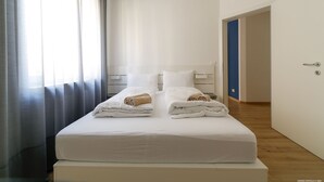 Uma cama de casal com roupa de cama branca e duas toalhas enroladas, piso de madeira e uma porta azul ao fundo.
