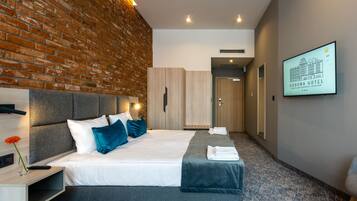 Quarto de hotel moderno com parede de tijolos, uma cama grande com almofadas azuis, um guarda-roupa de madeira e um quadro emoldurado na parede.