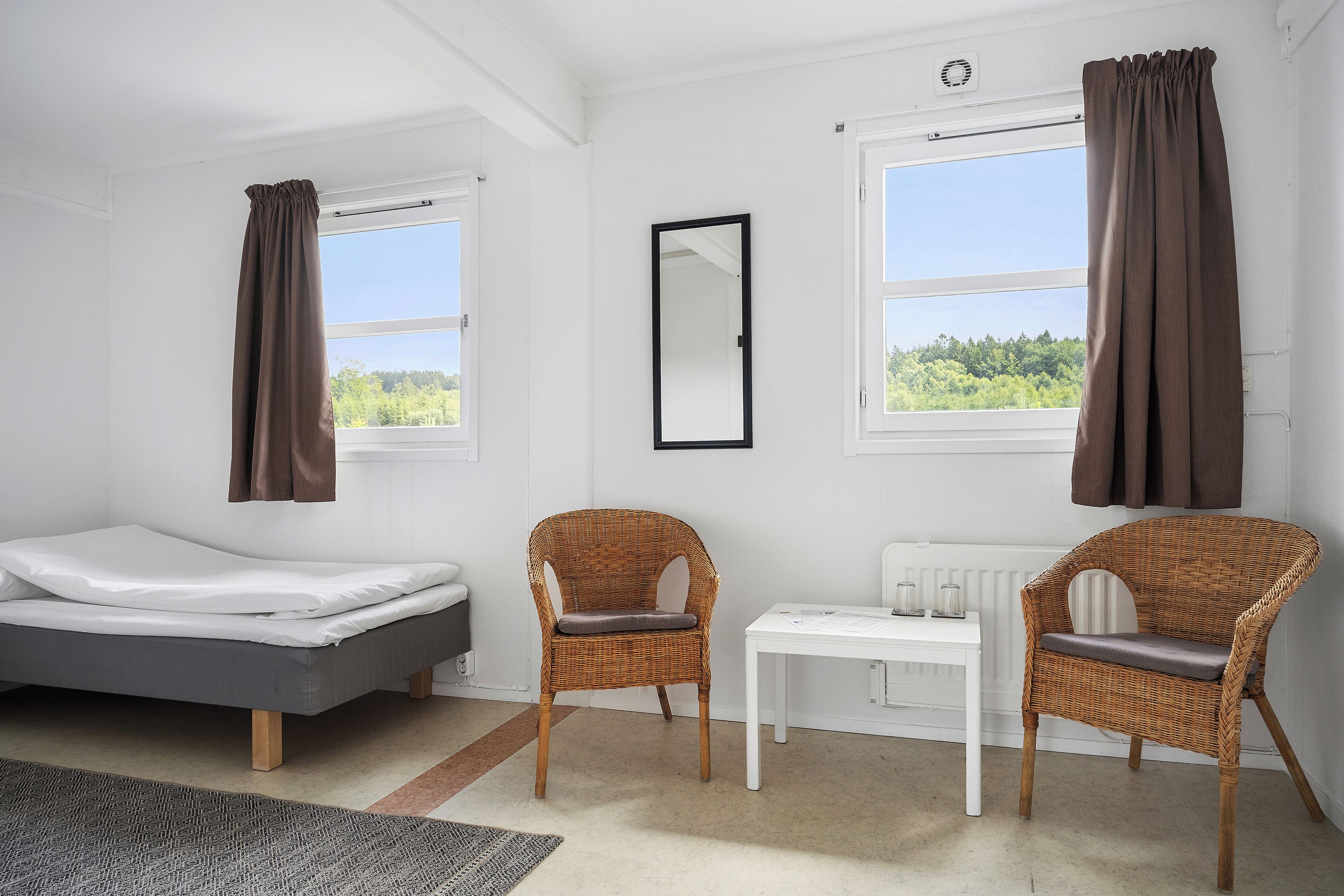 Um quarto com uma cama, duas cadeiras de vime, uma mesinha branca e duas janelas com cortinas.