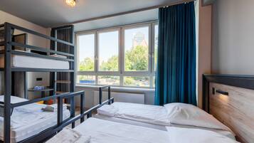 Quarto de hotel com beliches, uma janela grande com vista para prédios e árvores, e uma cortina azul.