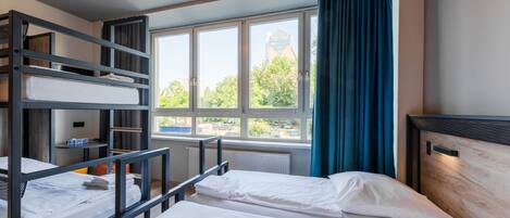 Quarto de hotel com beliches, uma janela grande com vista para prédios e árvores, e uma cortina azul.