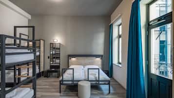 Quarto de hotel pequeno com uma cama de solteiro, uma cama individual, uma mesinha de cabeceira, um espelho e uma janela com cortinas.
