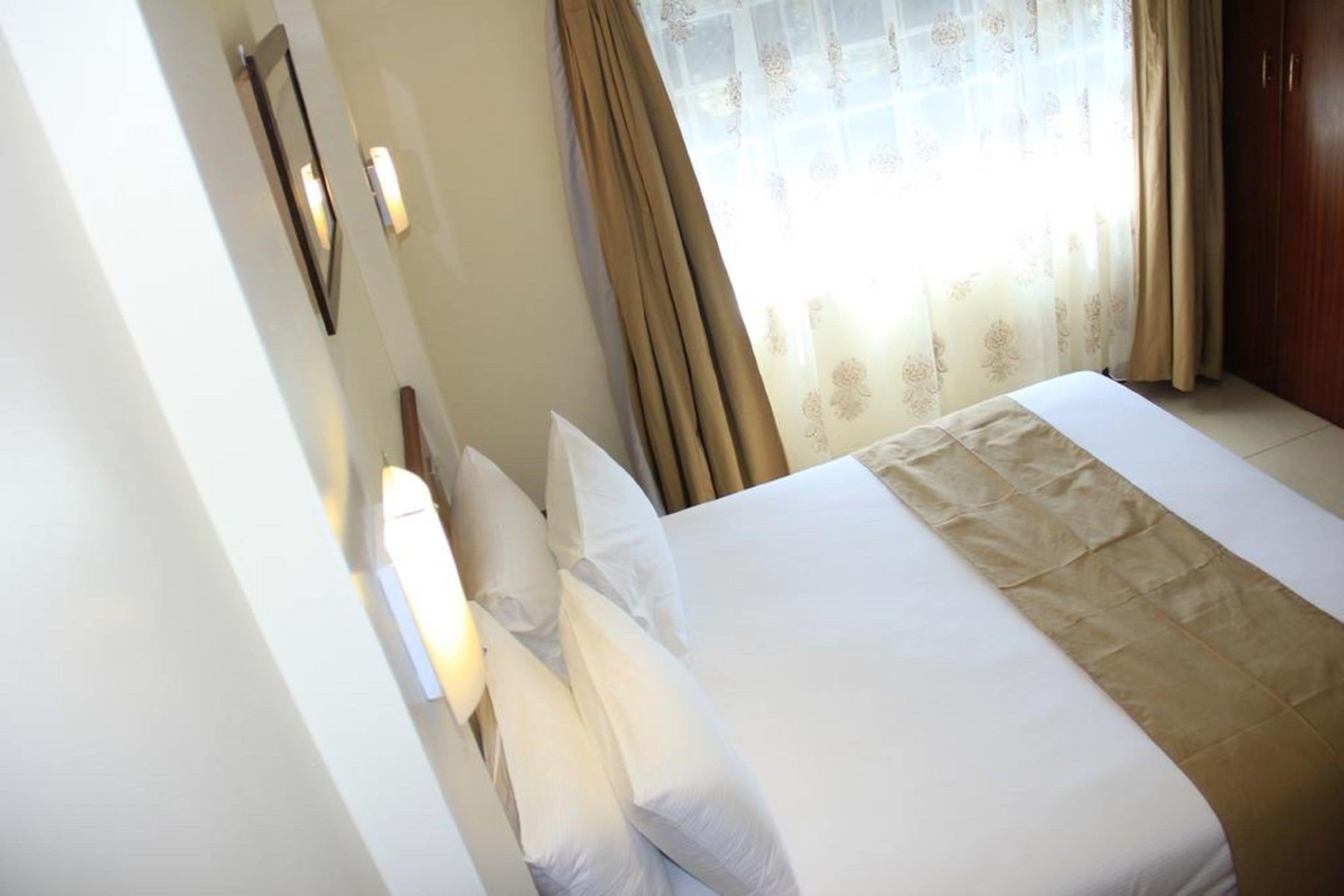 Quarto de hotel com cama, almofadas, mesa de cabeceira, luminária na parede e janela com cortinas.