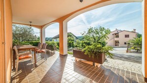 Alpendre coberto com mesa e cadeiras, plantas em vasos e vista para uma casa e paisagem.