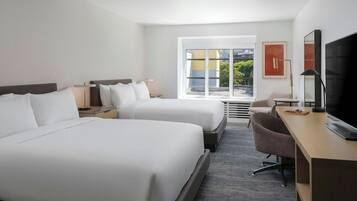 Quarto de hotel com duas camas, uma escrivaninha, uma cadeira, uma televisão e uma janela com vista para edifícios e árvores.