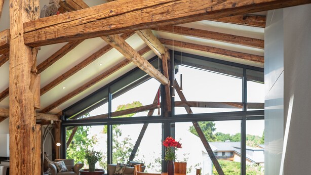 Um quarto espaçoso com teto alto, vigas de madeira, uma mesa de jantar, cadeiras, um sofá e grandes janelas com vista para o verde lá fora.