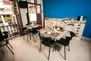 Uma área de refeições com uma mesa posta para dois, cadeiras, um balcão de buffet com comida e bebidas, e vista da cidade através de grandes janelas.