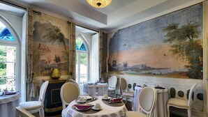 Uma sala de jantar com mesas redondas, cadeiras brancas e um grande quadro paisagístico na parede.
