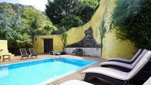 Piscina com espreguiçadeiras e uma pequena mesa, cercada por vegetação e um muro de pedra.