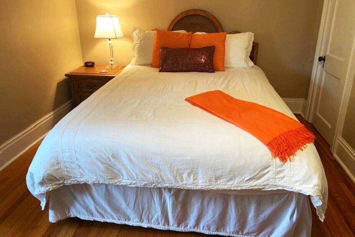 Cama arrumada com colcha branca, manta laranja e almofadas estampadas.