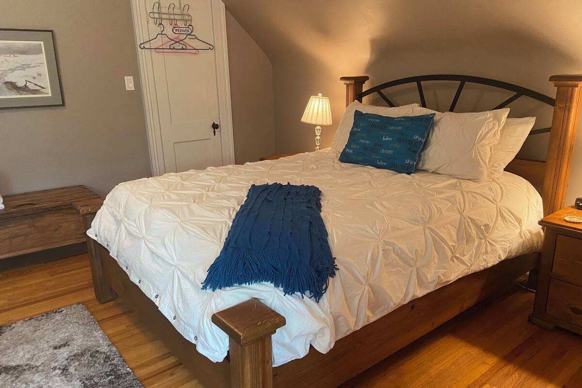 Quarto com cama, luminária de cabeceira, cabeceira de madeira, porta com cabideiro e quadro na parede.