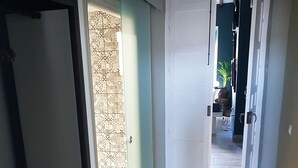 Um corredor com piso ladrilhado, uma porta que dá para um quarto com azulejos estampados e uma estante com toalhas dobradas.