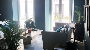 Uma sala de estar com um sofá, uma mesa de centro e um vaso de planta. Há duas janelas com cortinas, e um ventilador de teto é visível.