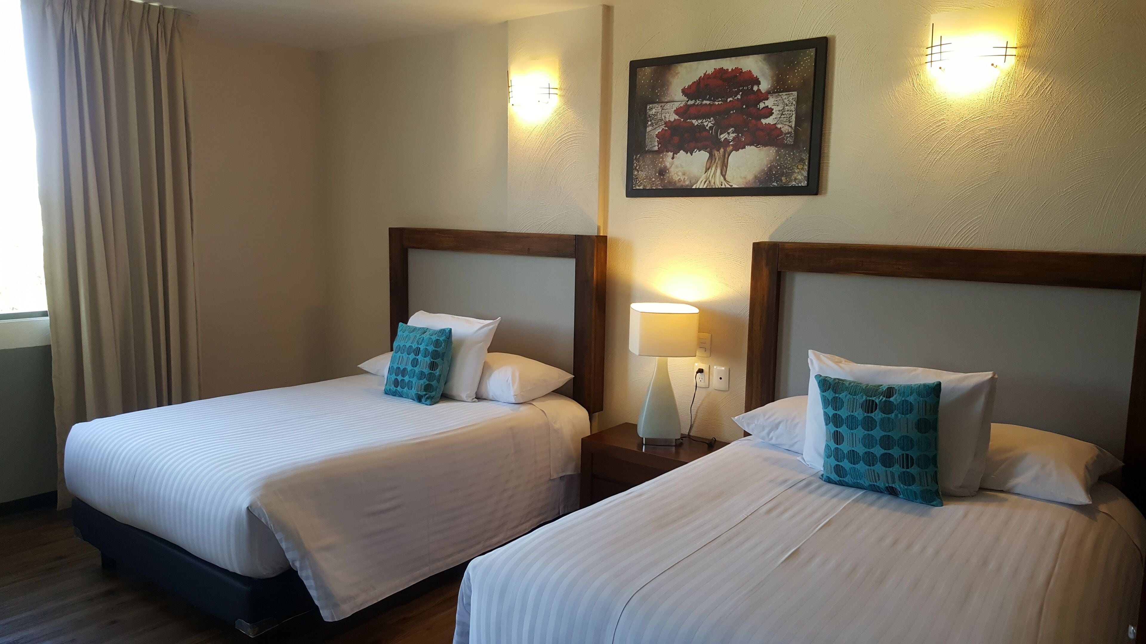 Quarto de hotel com duas camas, cada uma com cabeceira de madeira, roupa de cama branca e almofadas estampadas em tons de verde-água. H á um quadro na parede e uma mesa de cabeceira com abajur entre as camas.
