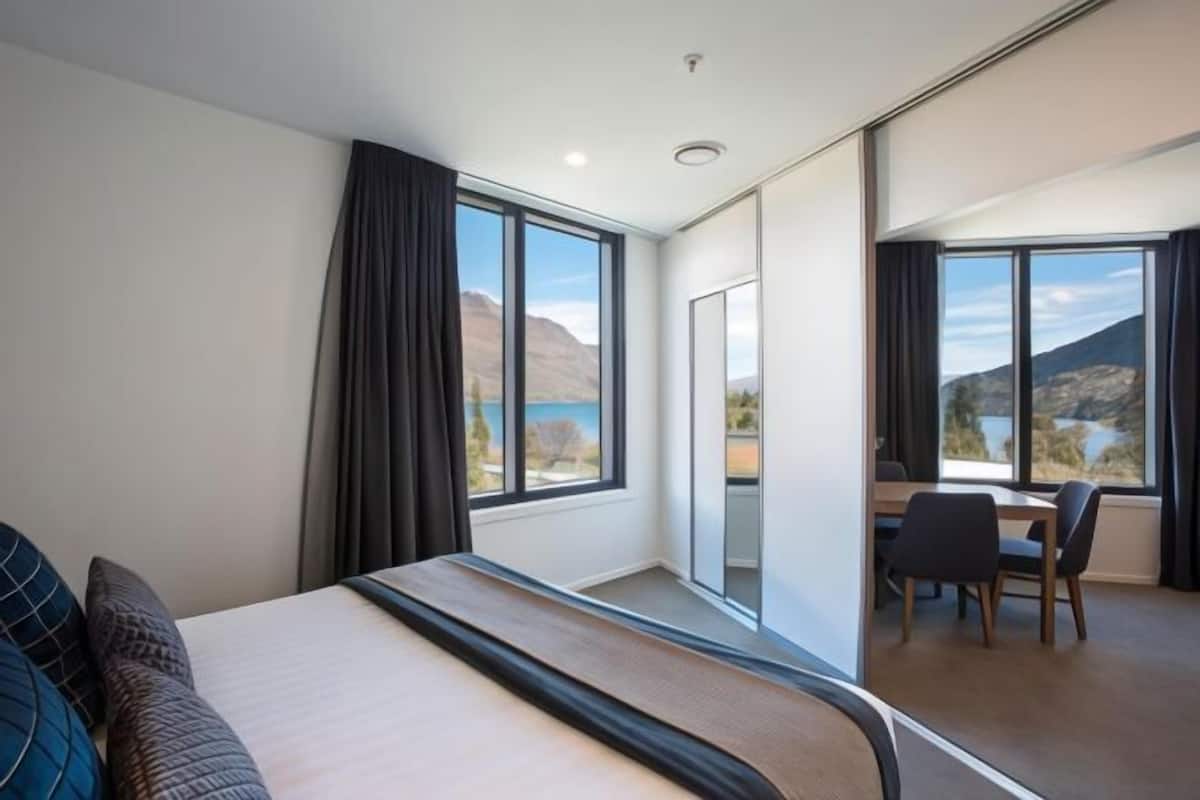 Quarto de hotel moderno com uma cama grande, uma mesa de trabalho e vista para montanhas pelas janelas.