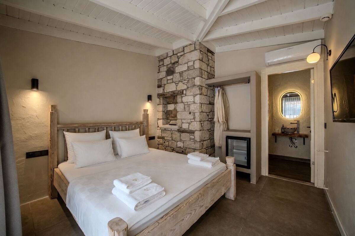 Quarto com lareira de pedra, cama com roupa branca, televisão pequena e banheiro com espelho e pia.