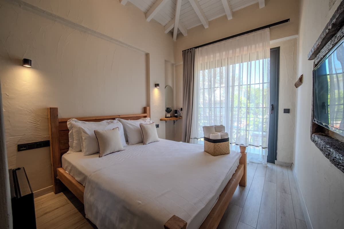Uma cama grande com cabeceira de madeira, uma mesa de cabeceira, uma luminária fixada na parede, uma janela com cortinas e vista para o verde lá fora.