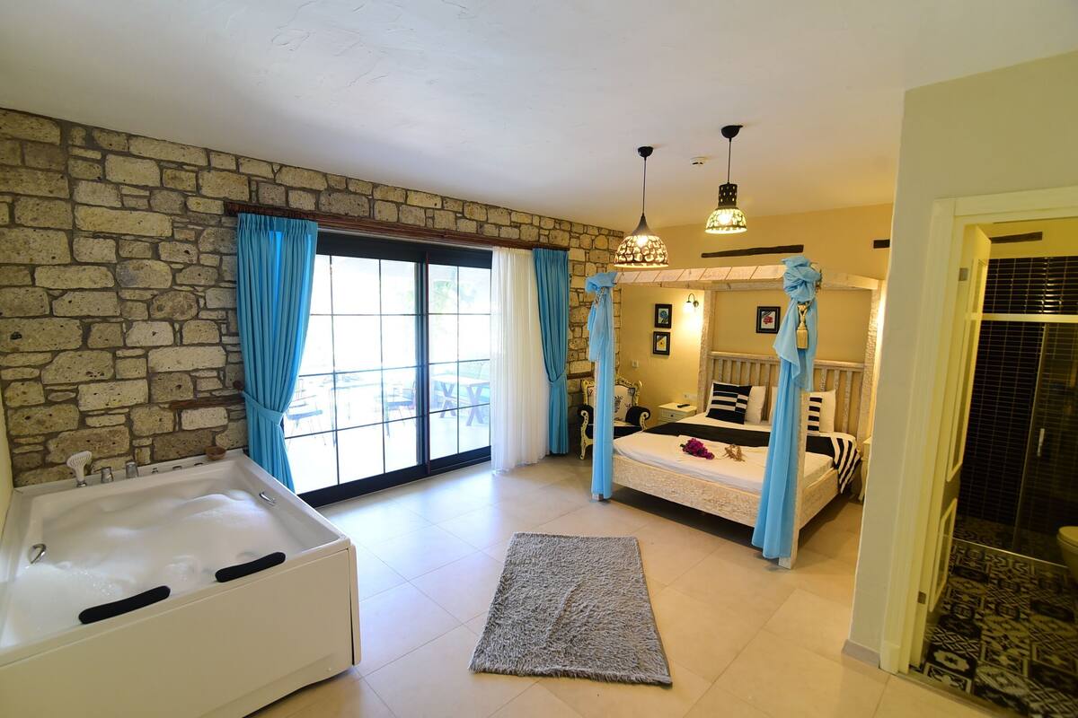 Quarto com parede de pedra, uma janela grande, cama com cortinas, banheira e área de chuveiro.