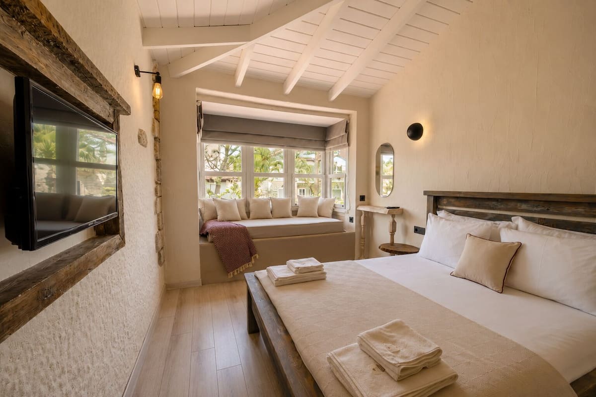 Quarto com cama, um banco com almofadas, televisão e janela com vista.