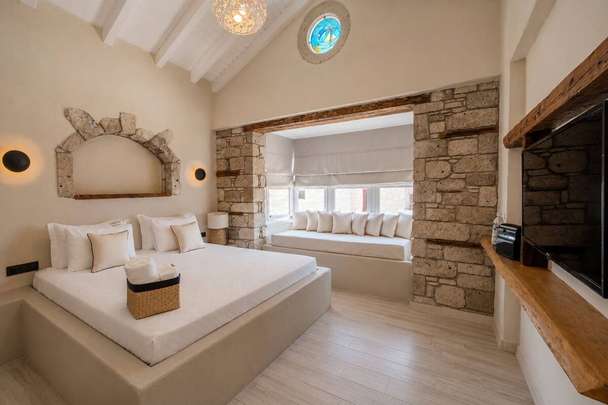 Um quarto com parede de pedra, uma cama com roupa de cama branca, um banco de pedra e uma janela com vista.