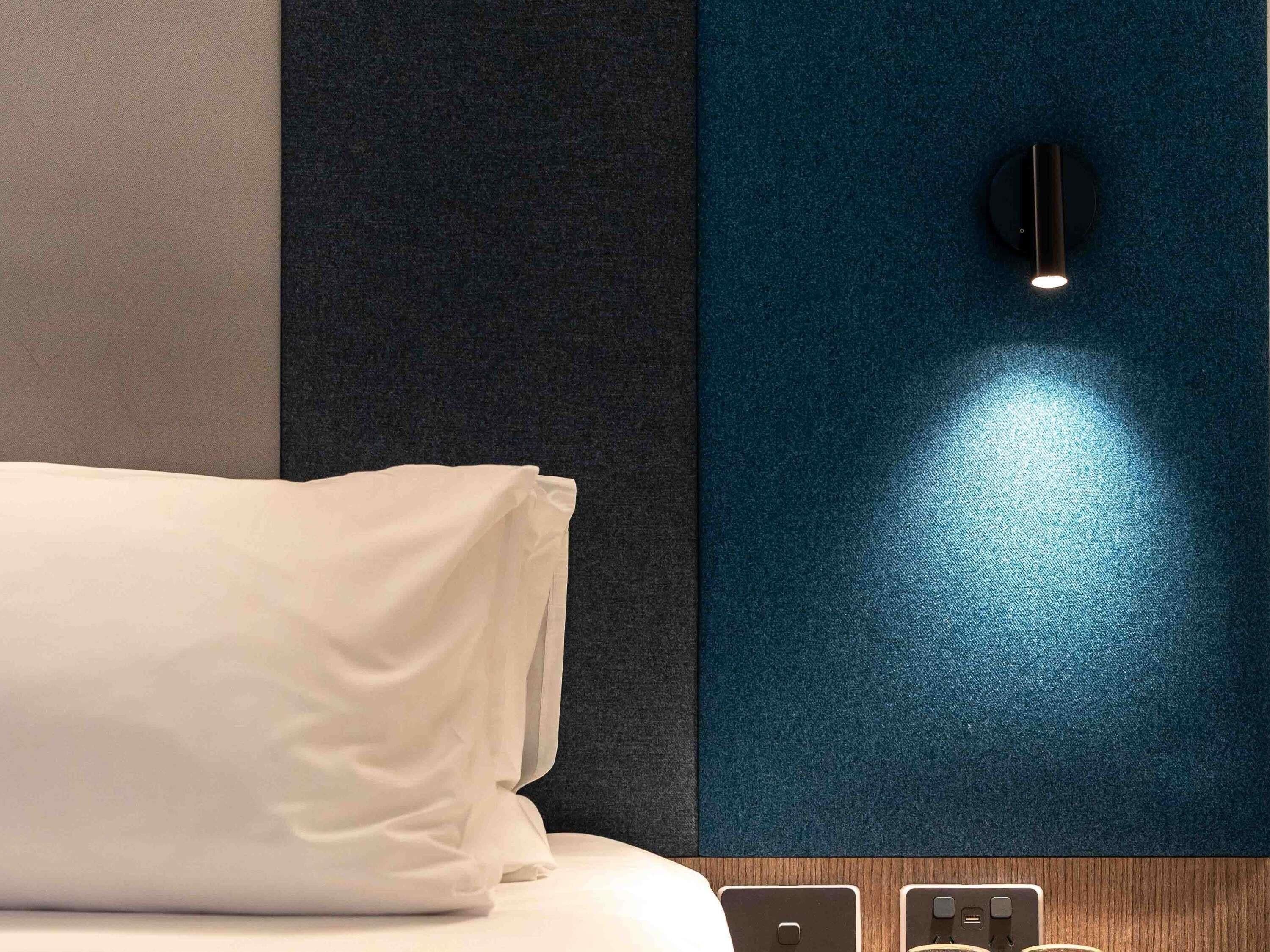 Quarto de hotel com uma cama, uma luminária fixada na parede e uma tomada.