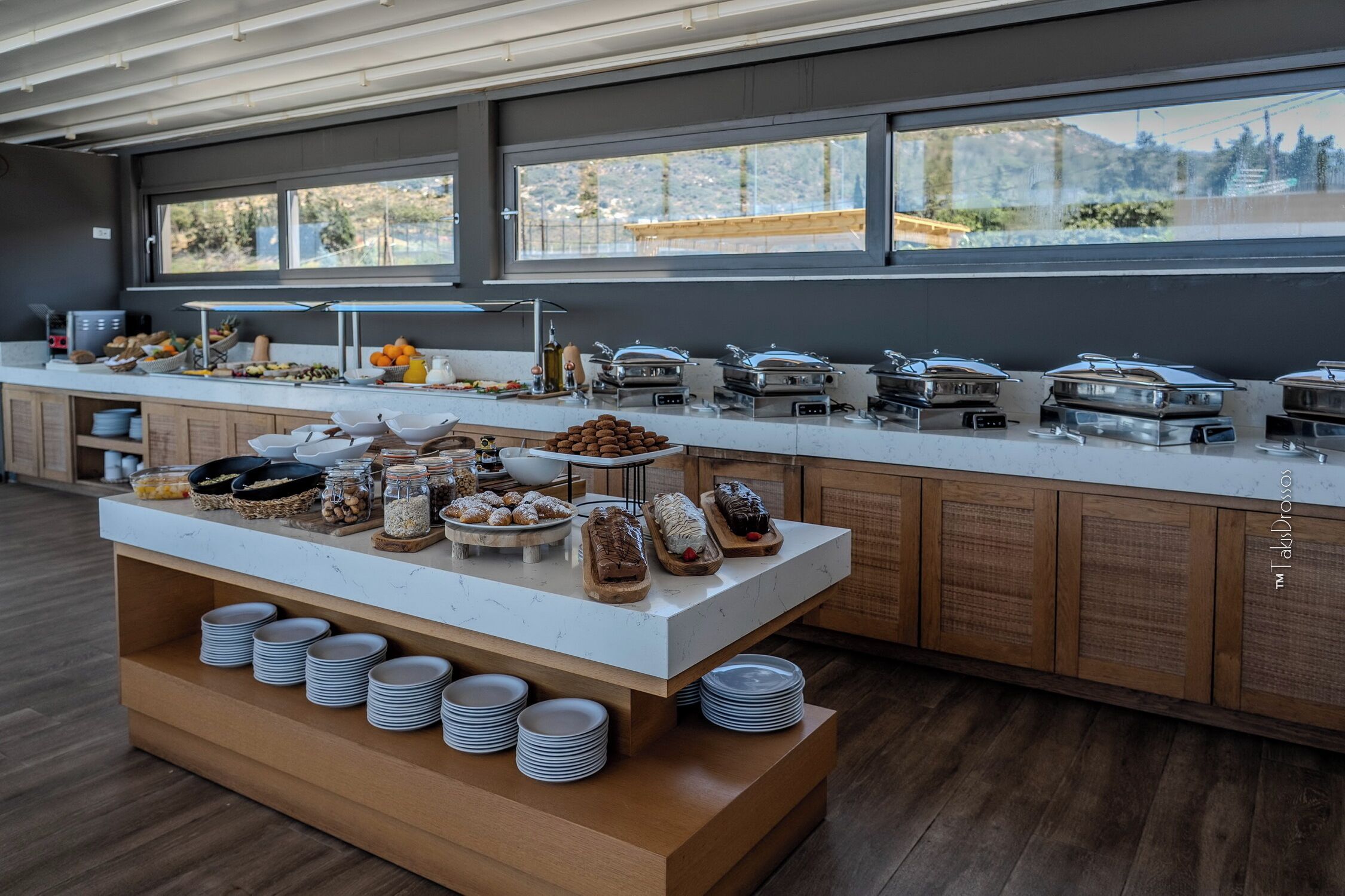 Disposição de buffet com diversos pratos e estações de cozinha, uma janela ampla com vista e design de cozinha moderno.