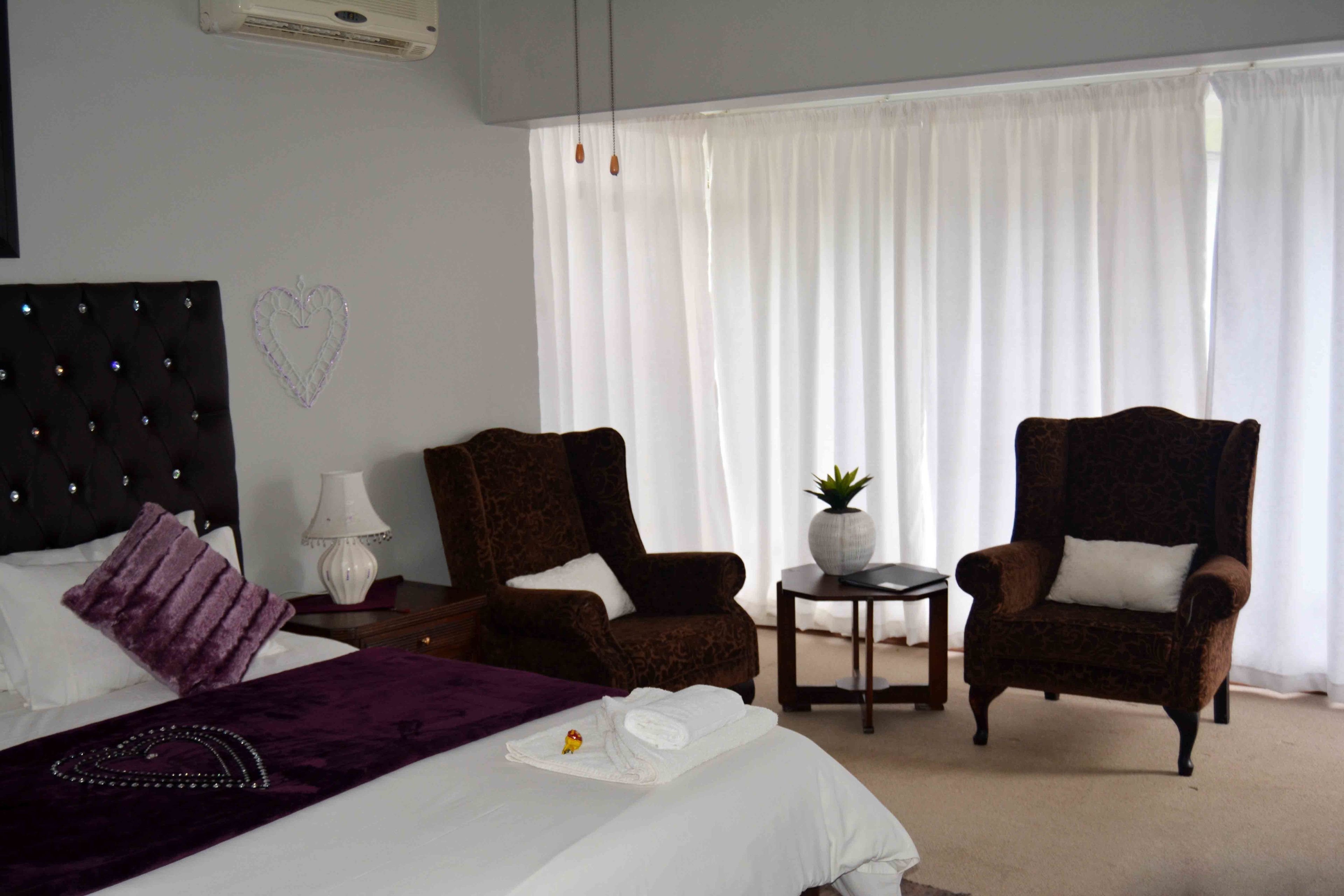 Quarto de hotel com uma cama grande, duas poltronas, uma mesinha de centro com uma planta e uma cortina branca.