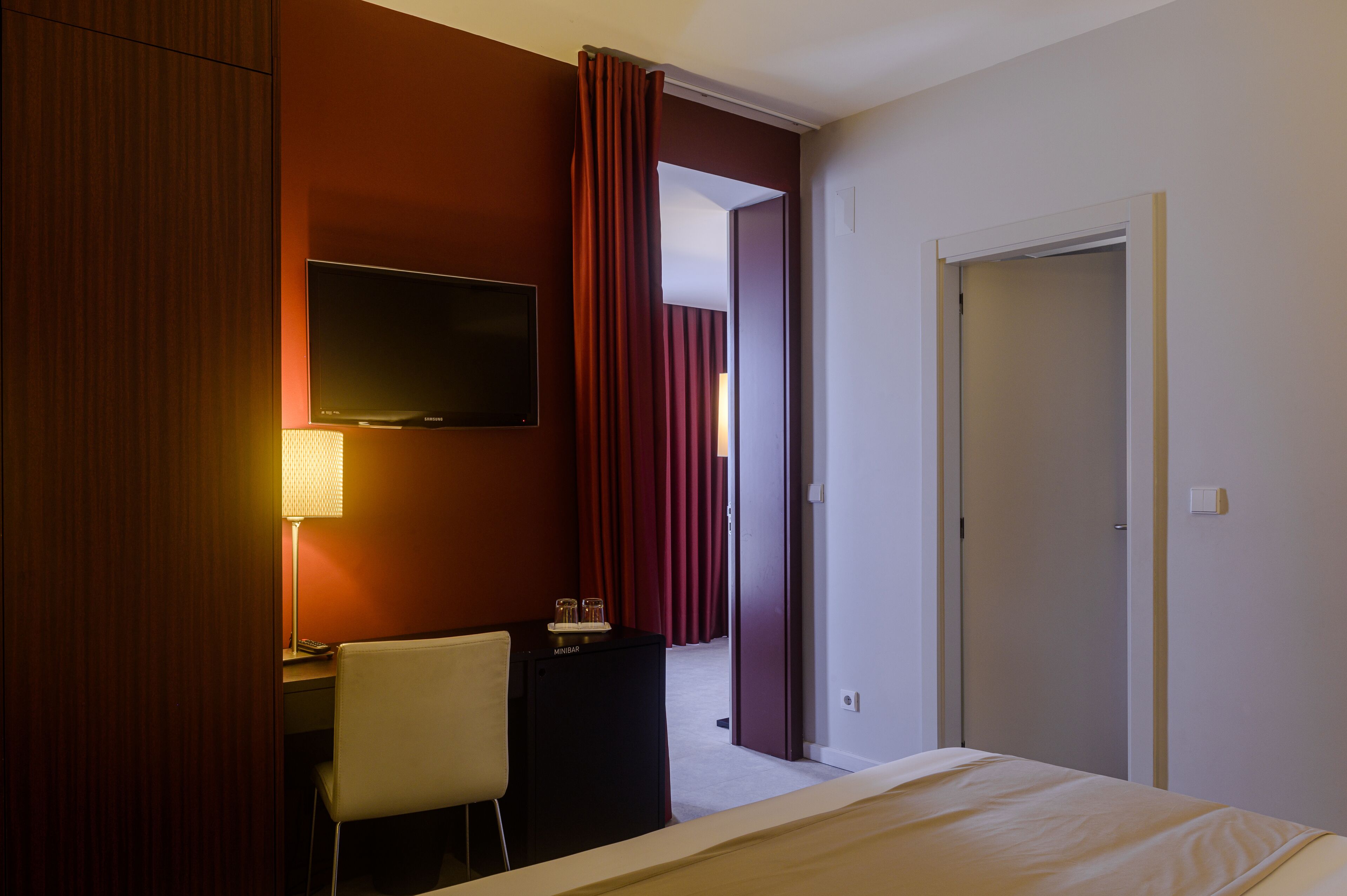 Quarto de hotel com cama, escrivaninha, cadeira, televisão e porta.