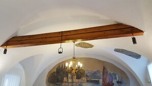 Quarto com teto de vigas de madeira, um candelabro e um mural com um castelo e veleiros.