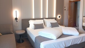 Quarto de hotel moderno com uma cama grande, mesas de cabeceira, um banquinho redondo e uma porta de madeira.