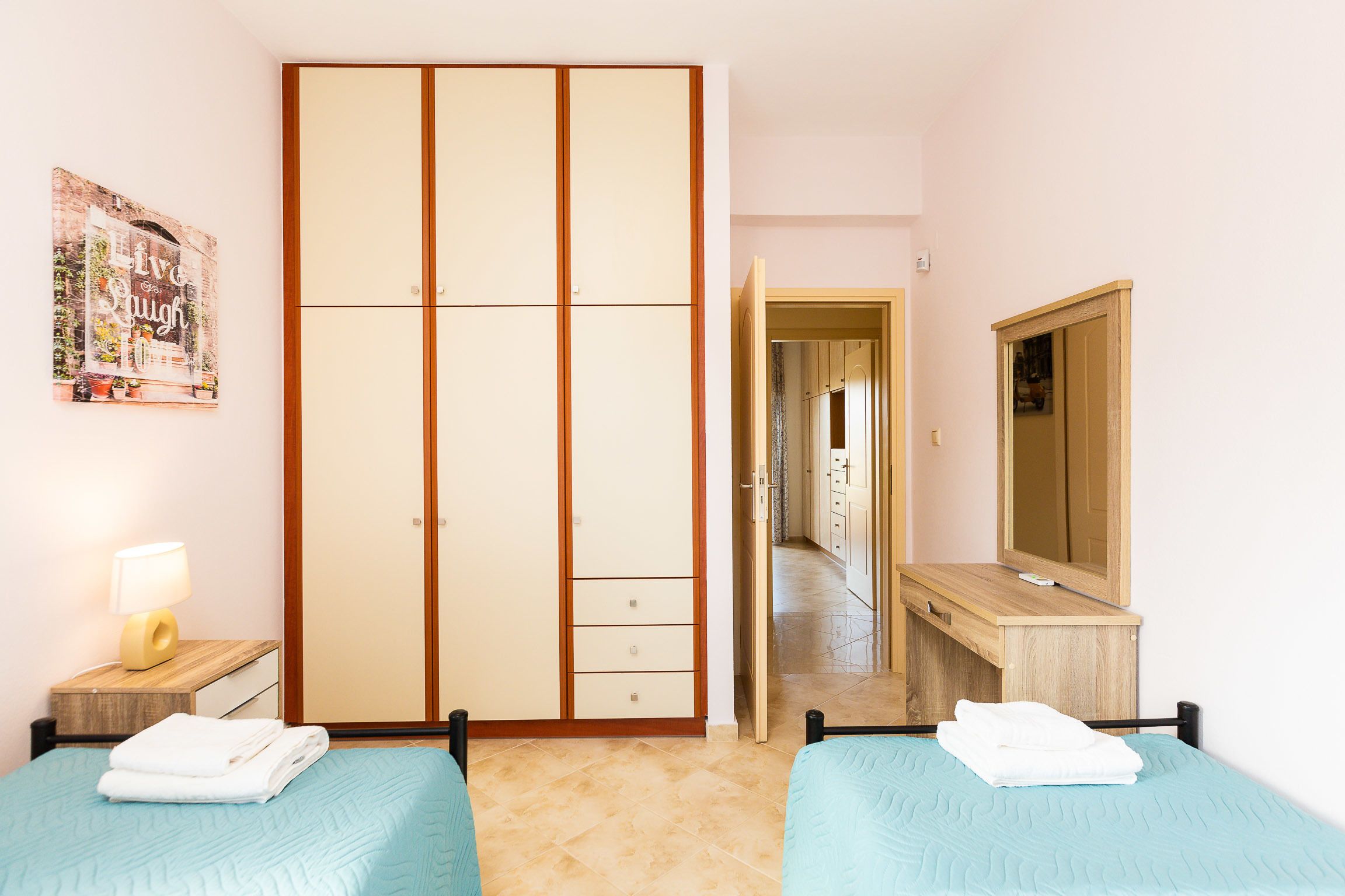 Quarto de hotel com duas camas, um guarda-roupa, uma mesa de cabeceira com abajur, um espelho e um banheiro visível através de uma porta aberta.