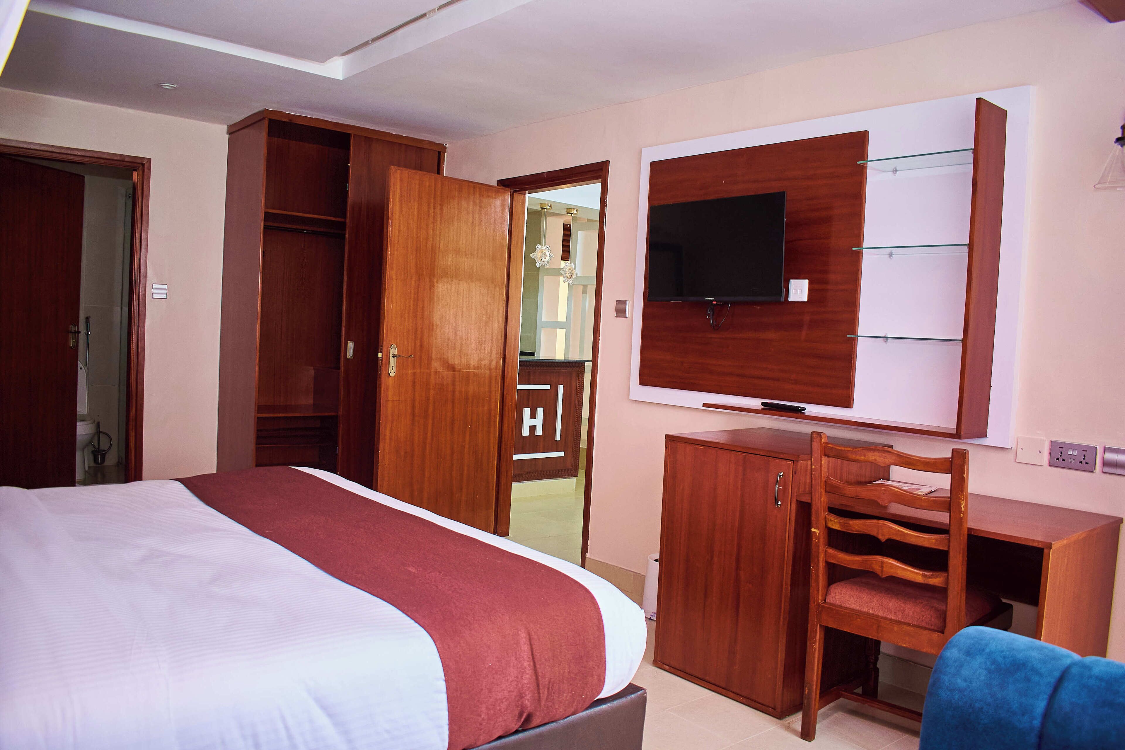 Quarto de hotel com cama, escrivaninha, cadeira, guarda-roupa e televisão na parede.