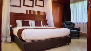 Uma cama grande com cabeceira de madeira, uma poltrona de couro preto e uma janela com cortinas leves.