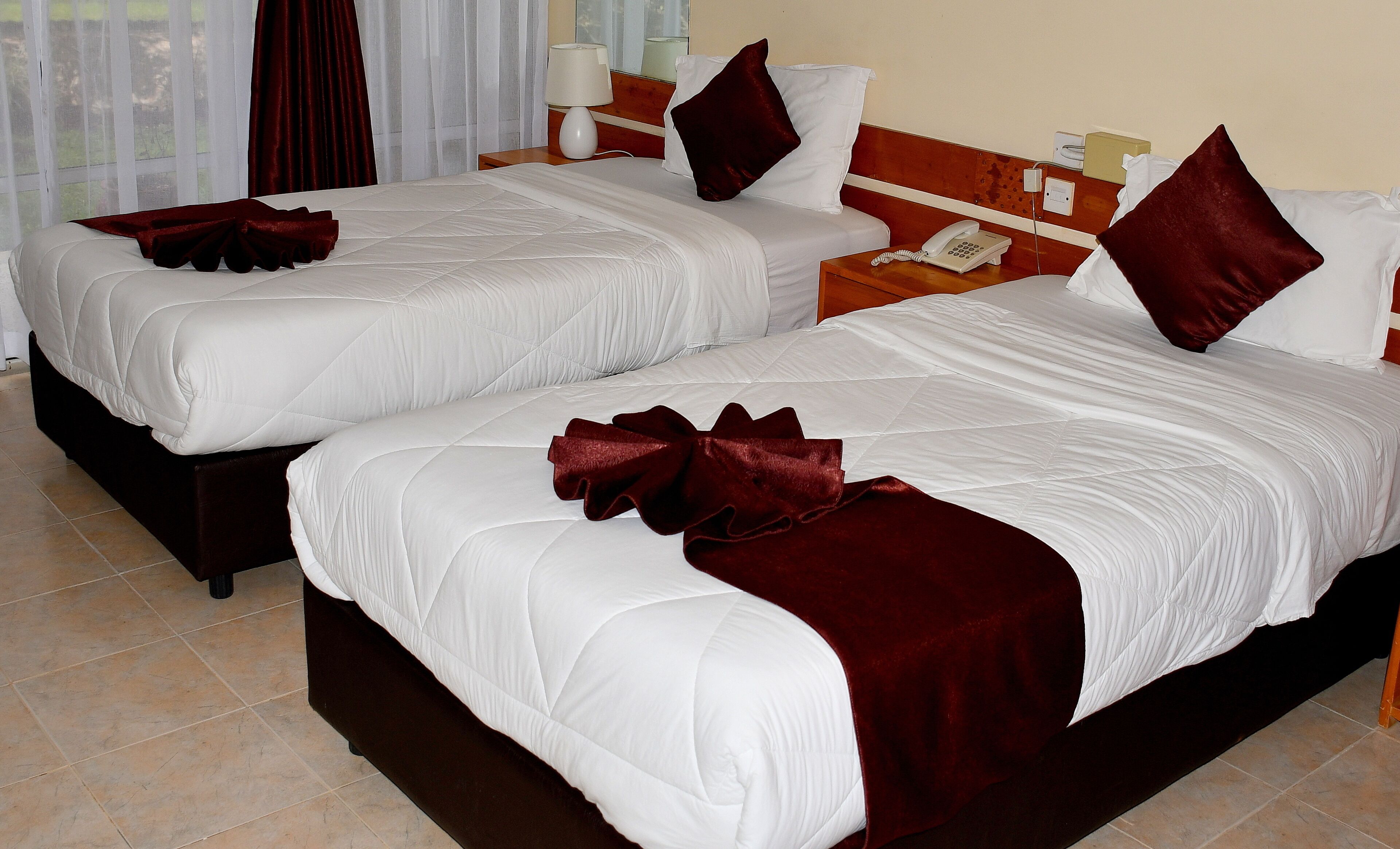Duas camas de casal com roupa de cama branca e detalhes em vermelho escuro, um telefone ao lado da cama e uma janela com cortinas.