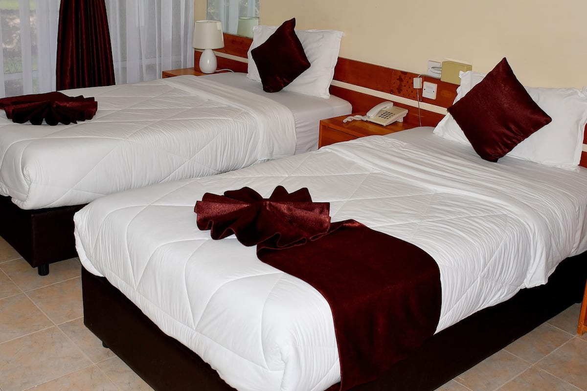 Duas camas de casal com roupa de cama branca e detalhes em vermelho escuro, um telefone ao lado da cama e uma janela com cortinas.