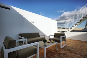 Terraço no telhado com parede branca, dois sofás, uma mesa com planta e uma escadaria.