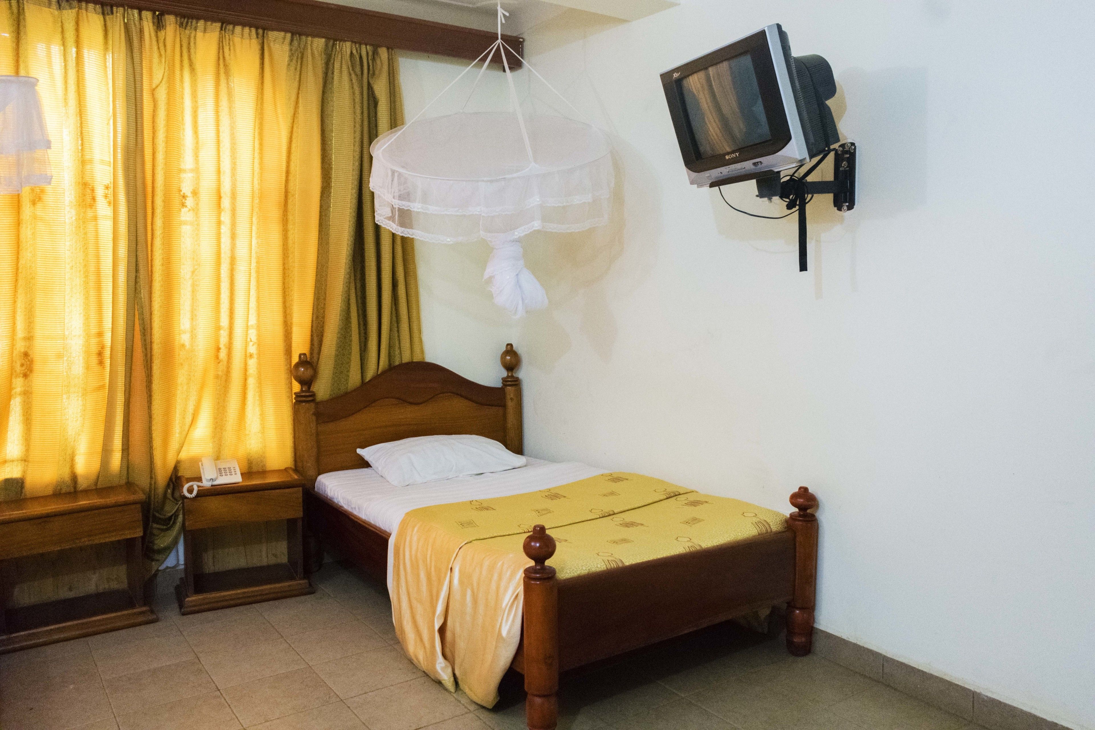 Quarto com cama de madeira, mesa de cabeceira com telefone, mosquiteiro suspenso e televisão na parede.