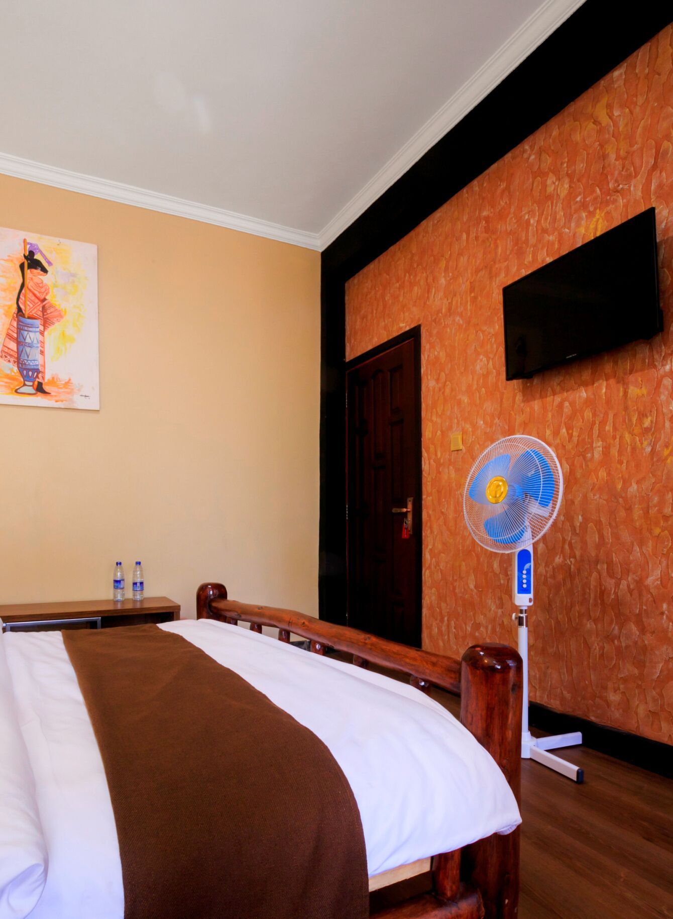 Quarto de hotel com uma cama grande, uma escrivaninha de madeira, TV fixada na parede e um ventilador.