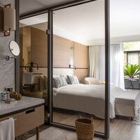 Quarto de hotel moderno com divisória de vidro, cama, sofá, mesa pequena, abajur e banheiro com espelho e amenities.