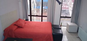 Quarto de hotel com cama, uma pequena mesa, um banco e janela com vista para a rua.
