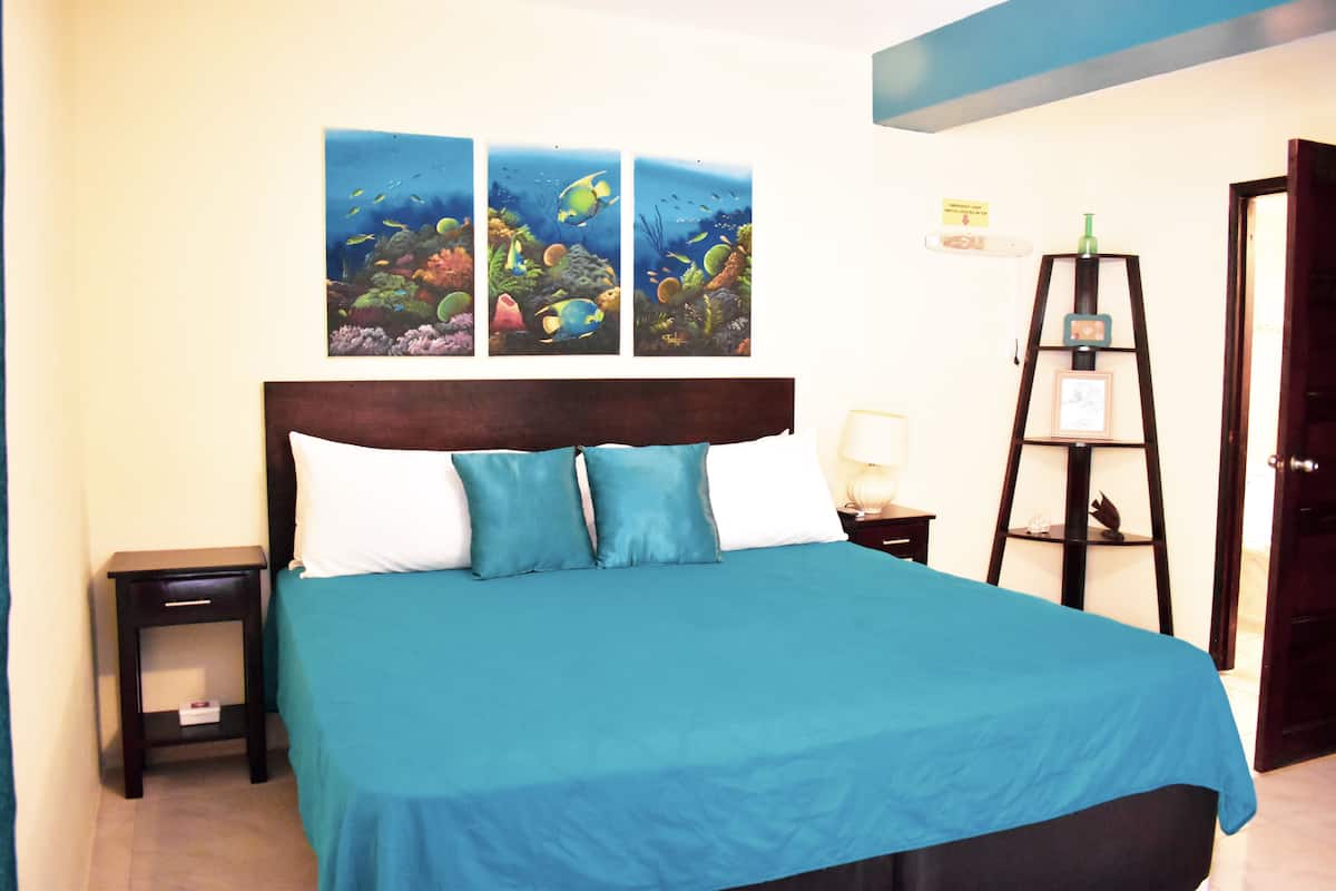 Uma cama com colcha azul-turquesa e travesseiros brancos, uma mesa de cabeceira com abajur, e três quadros com pinturas subaquáticas na parede.