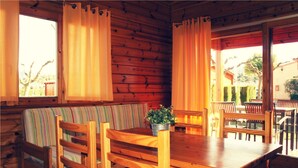 Interior de uma cabana de madeira com uma mesa de jantar, cadeiras, um banco e uma janela com cortinas.