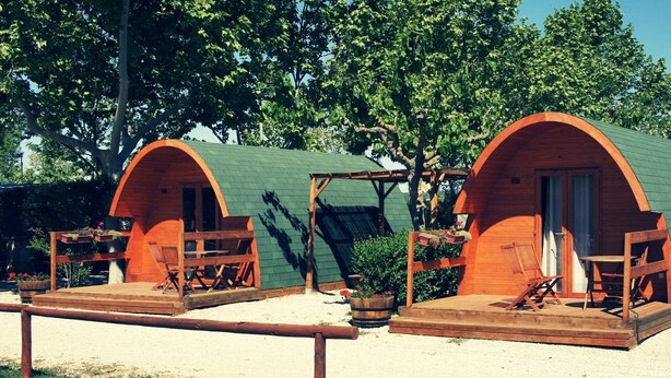 Duas cabanas de madeira com telhados verdes, cada uma com varanda e móveis de madeira, além de vasos com plantas.