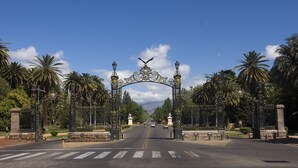 Uma entrada imponente com portões ornamentados e estátuas, ladeada por palmeiras e um céu claro.