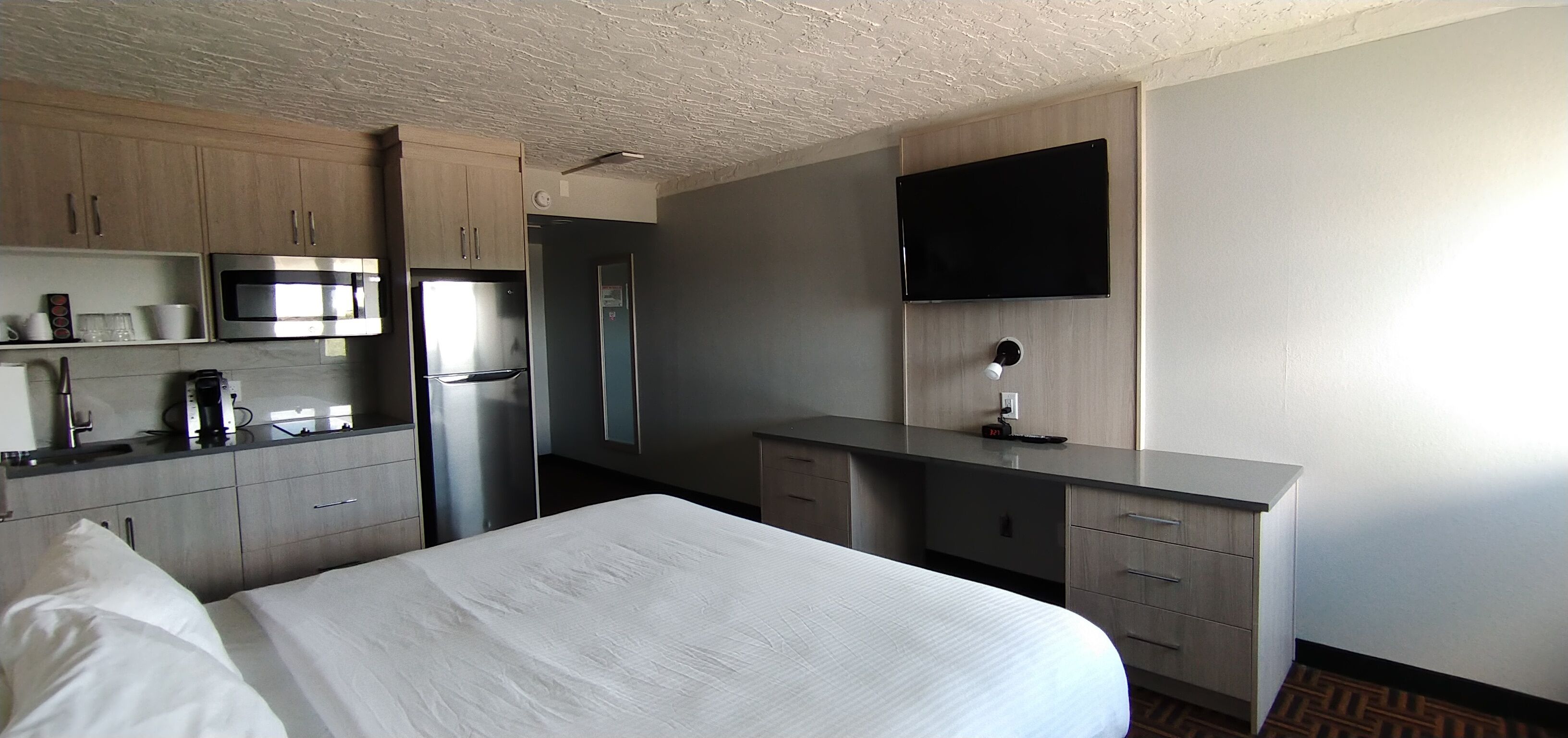 Quarto de hotel com uma cama, cozinha compacta com micro-ondas, geladeira e cafeteira, televisão de tela plana na parede e uma pequena escrivaninha com gavetas.