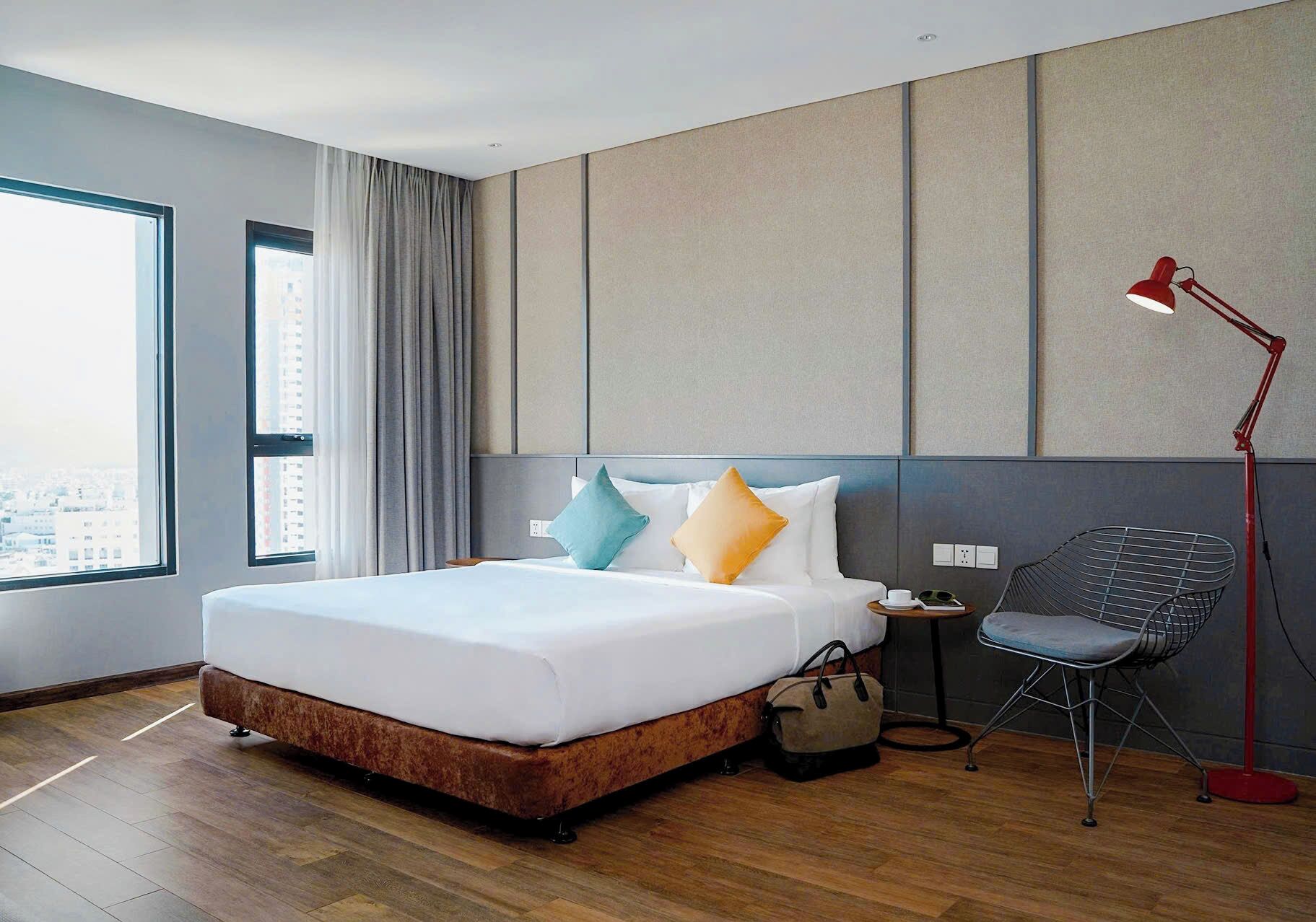 Quarto de hotel com uma cama grande, uma cadeira, uma escrivaninha e uma janela com cortinas.