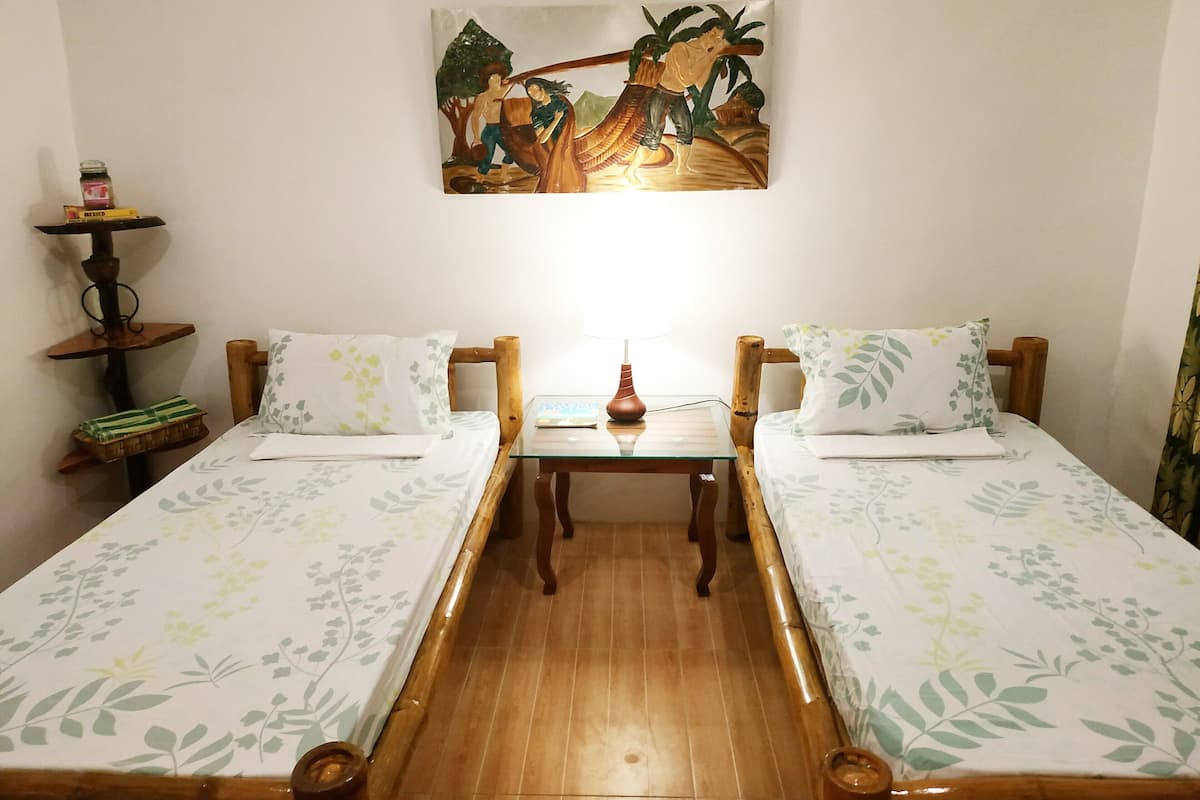 Quarto com duas camas de solteiro, uma mesa pequena com abajur, uma estante com um pote e um quadro na parede.