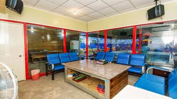 Quarto com uma mesa central, cadeiras azuis, um ventilador e uma janela com vista para uma piscina.