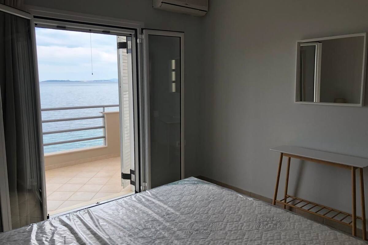 Quarto com cama, uma pequena mesa, espelho e porta de vidro deslizante que dá para uma varanda com vista para o mar.